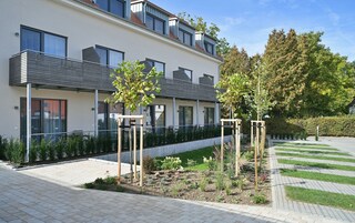 Um edifício moderno com varanda, um jardim com árvores jovens e um caminho pavimentado.