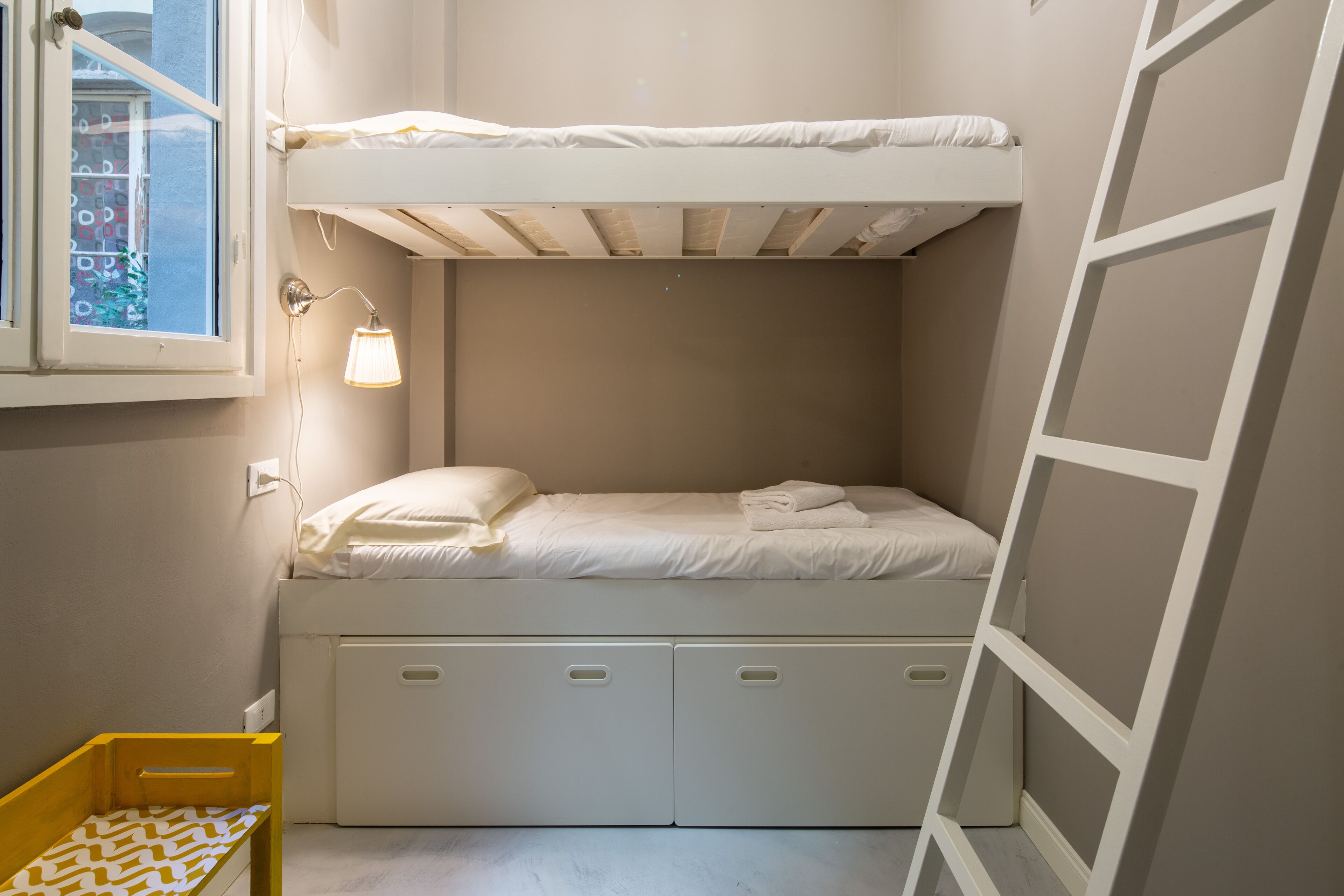 Uma cama de solteiro com uma escrivaninha embaixo, uma luminária de cabeceira e uma janela com vista para o exterior.