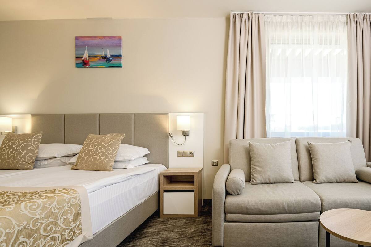 Quarto de hotel com cama, sofá, mesa de cabeceira, pintura e janela com cortinas.