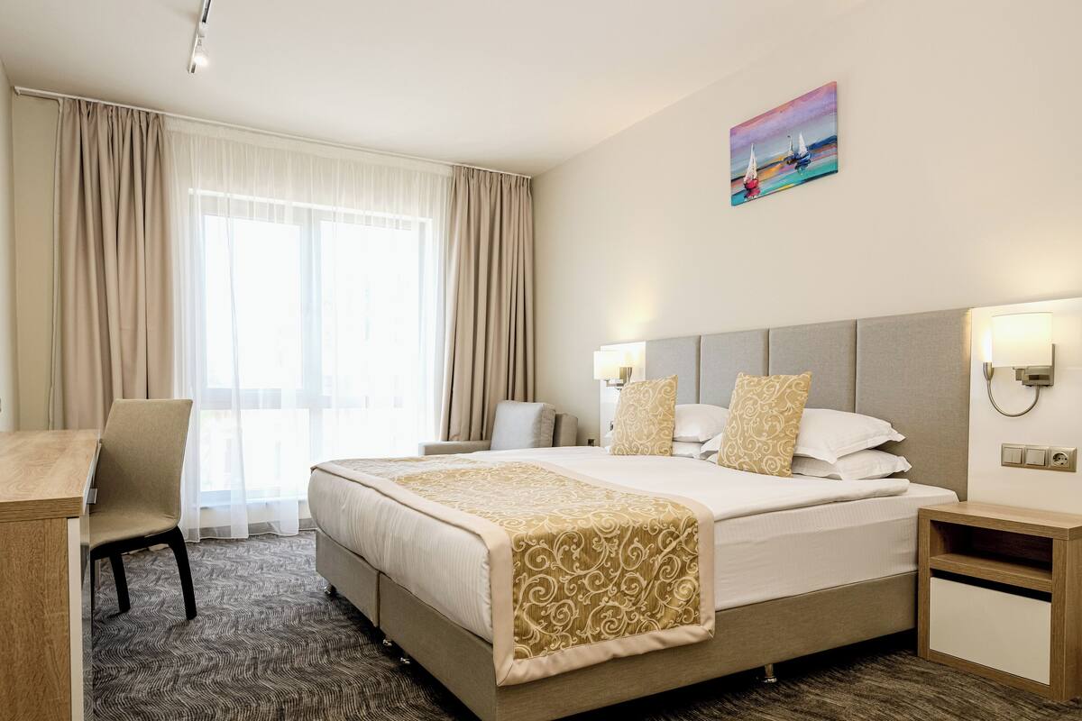 Quarto de hotel com cama grande, escrivaninha, cadeira e mesa de cabeceira.