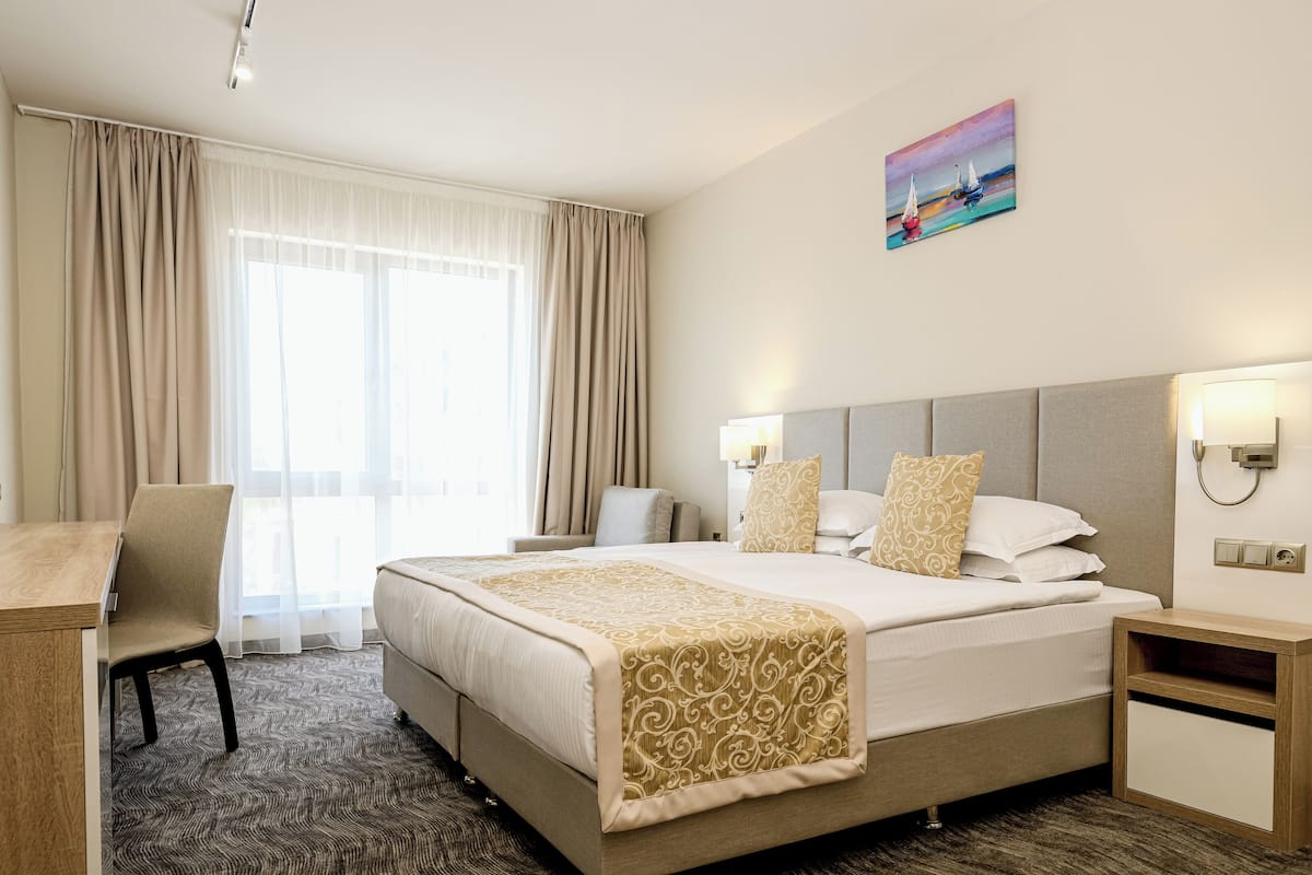 Quarto de hotel com cama grande, escrivaninha, cadeira e mesa de cabeceira.