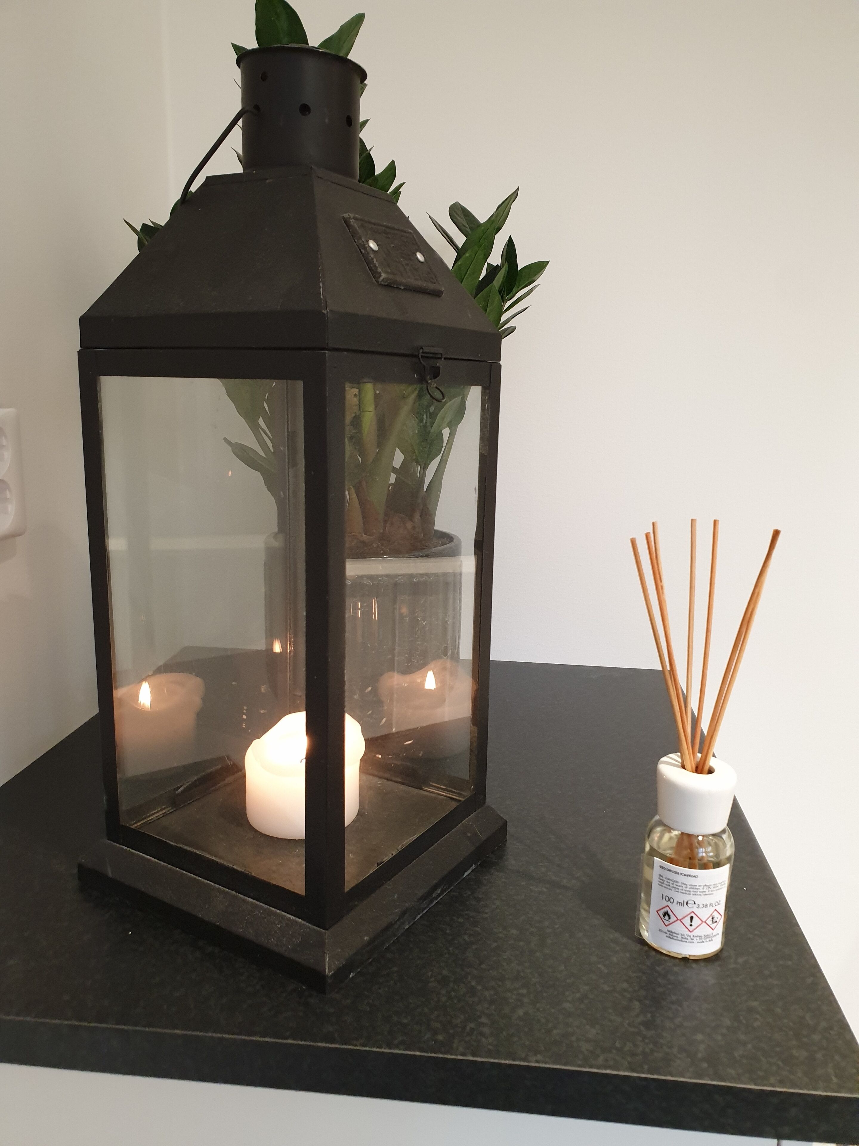 Um candeeiro preto com uma vela no interior, colocado sobre uma bancada escura ao lado de um pequeno difusor de vime.