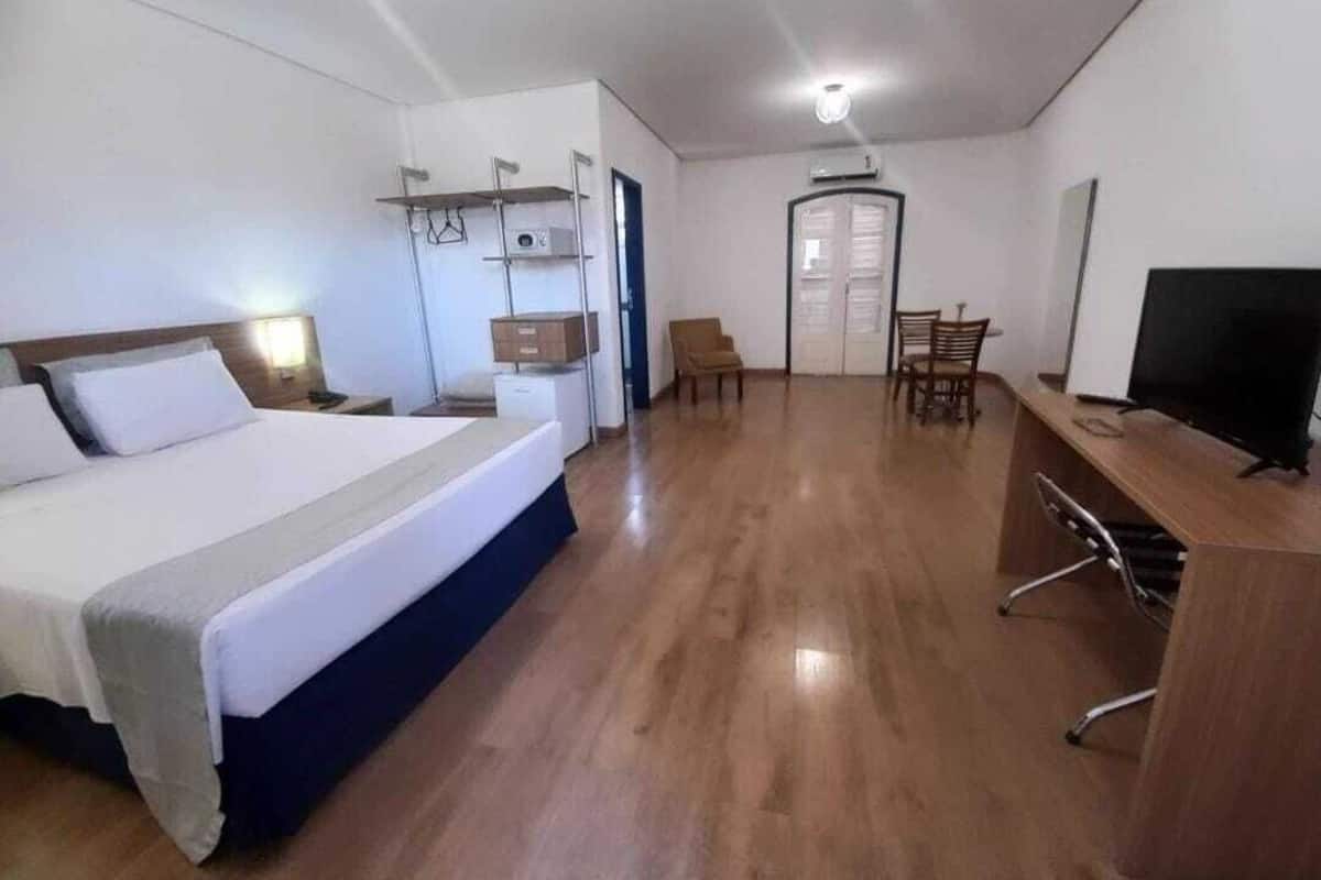 Quarto de hotel com cama, escrivaninha, cadeira, televisão e guarda-roupa.