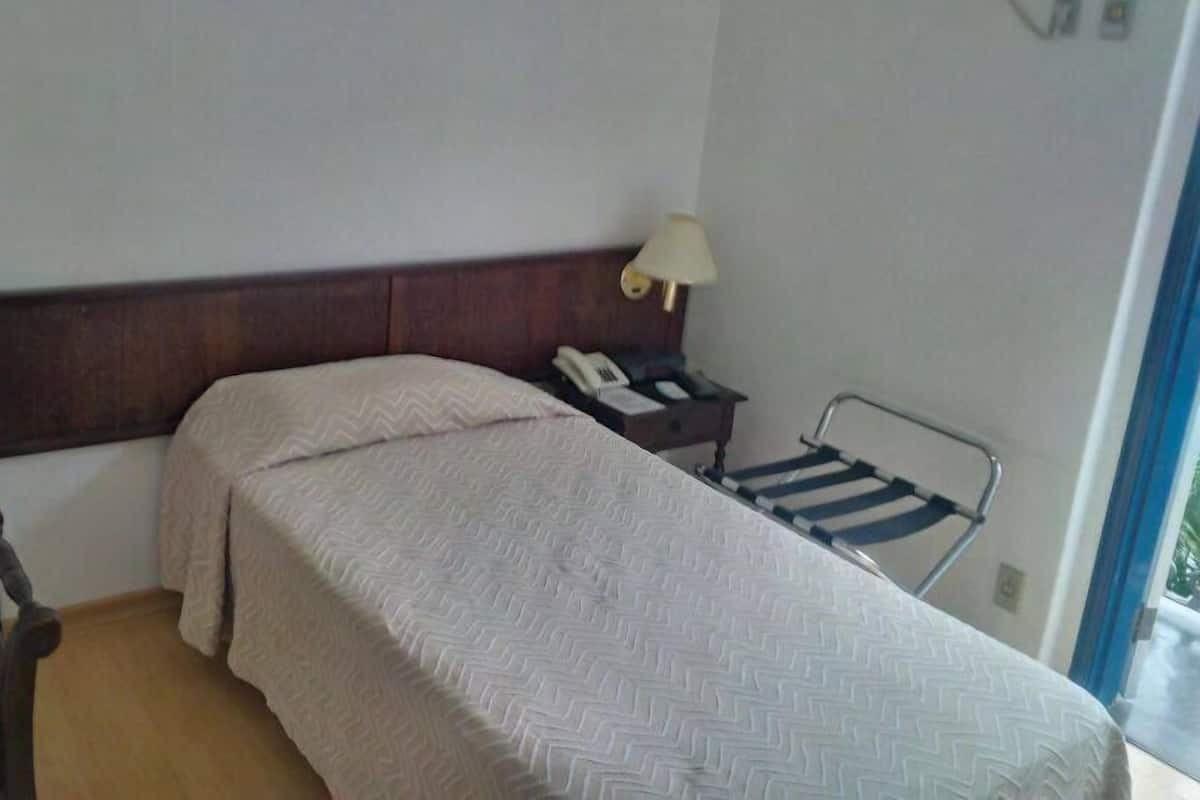 Uma cama de solteiro com colcha estampada, uma mesa de cabeceira com abajur, uma cadeira e uma janela com vista.