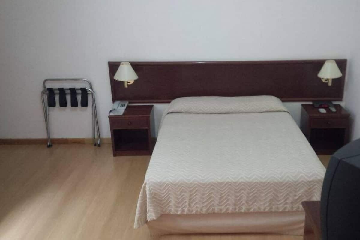 Quarto de hotel com uma cama, duas mesas de cabeceira, uma lâmpada em cada mesa, uma cadeira e uma mala.