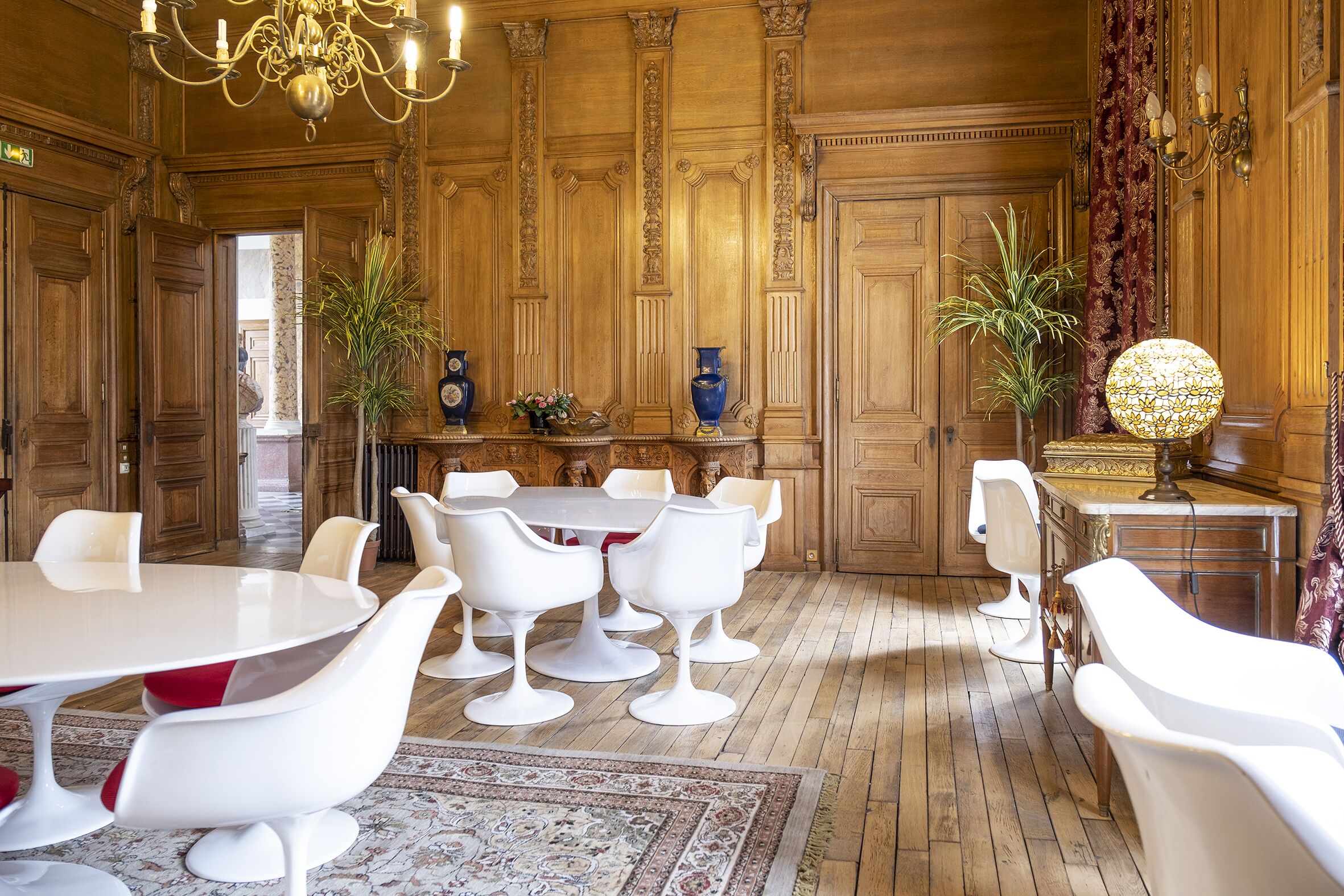Uma sala de jantar com uma mesa redonda e cadeiras brancas, cercada por painéis de madeira e um candelabro.
