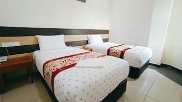 Quarto de hotel com duas camas, cada uma com cabeceira de madeira, roupa de cama branca com padrões vermelhos e dourados, e uma pequena mesa de cabeceira com uma luminária.