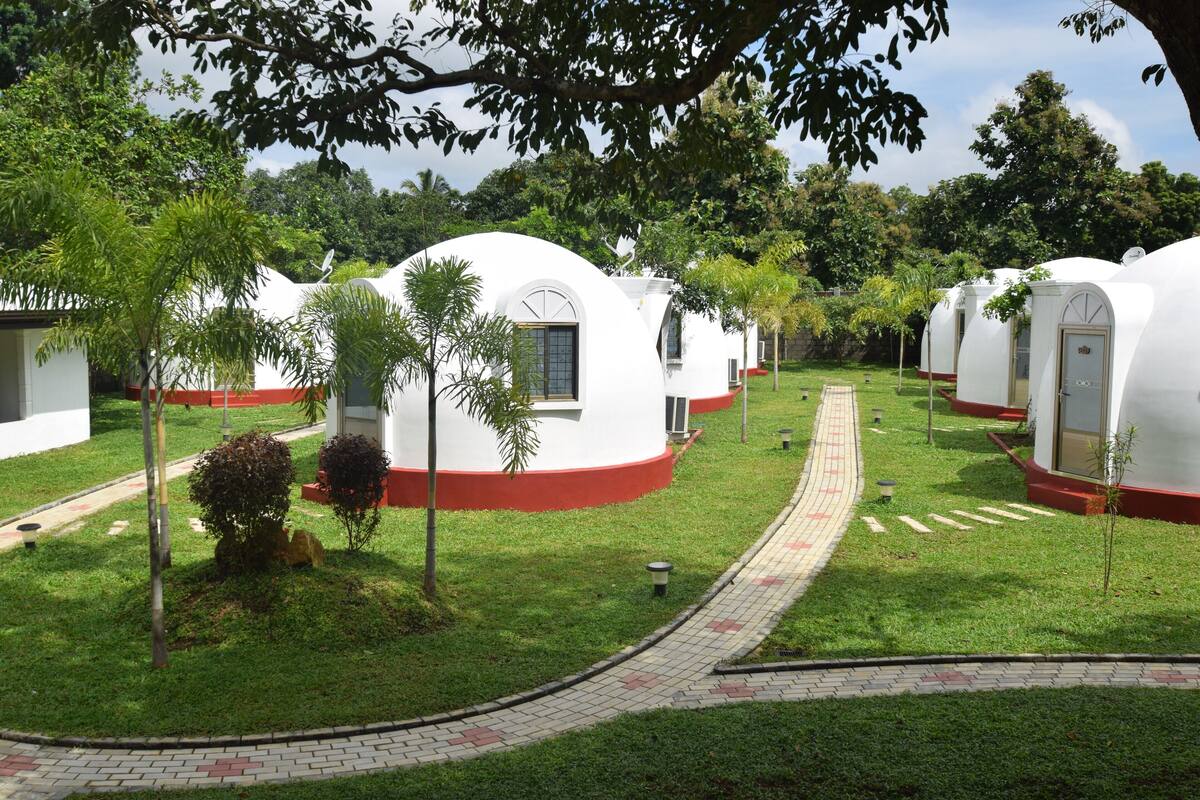 Uma fileira de cabanas brancas em formato de cúpula, com detalhes vermelhos, cercada por grama bem cuidada e árvores.