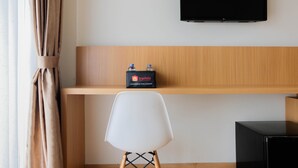 Quarto de hotel com escrivaninha de madeira, cadeira branca, TV preta fixada na parede e prateleira com um produto travelio.