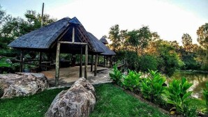 Um pavilhão com telhado de palha e estrutura de madeira, rodeado por vegetação exuberante e um lago.