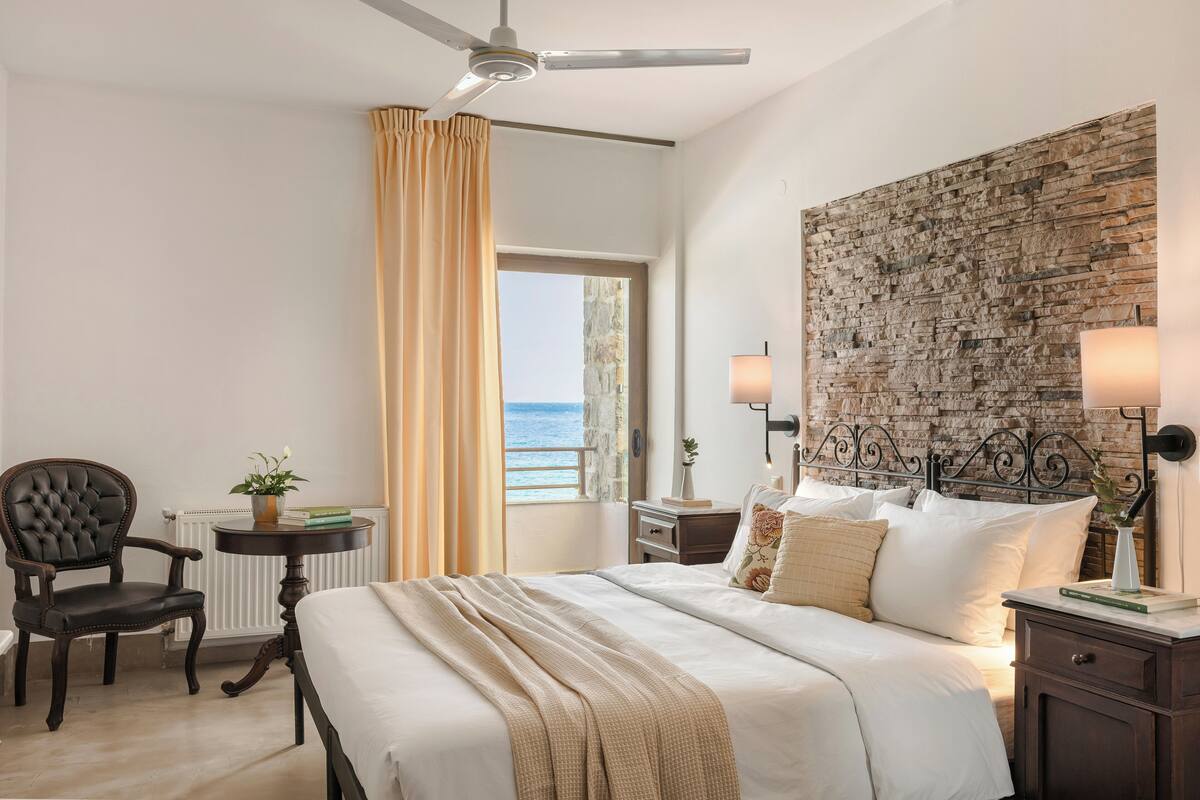 Quarto de hotel com uma cama grande, uma cadeira, uma mesinha com uma planta, uma mesa de cabeceira com abajur e vista para o mar pela janela.
