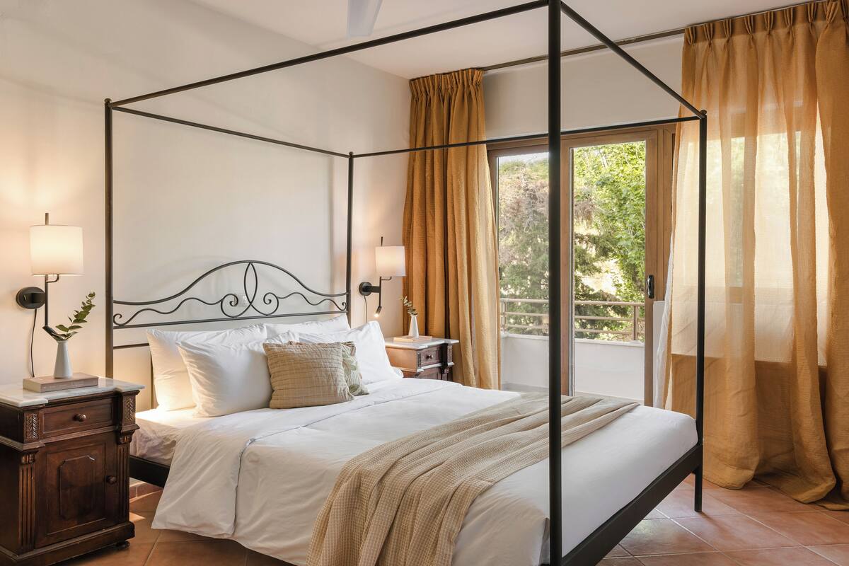 Uma cama de dossel com cabeceira, uma mesa de cabeceira com abajur e vista para o exterior por uma porta de correr.