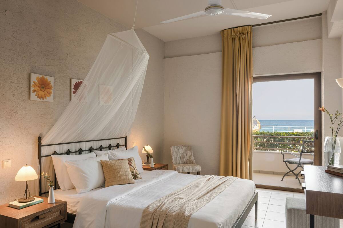 Quarto com cama, mesas de cabeceira, um banquinho e vista para o mar.