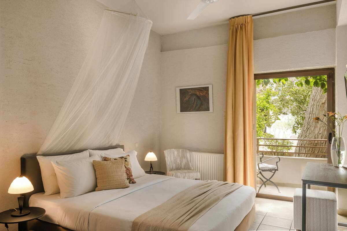 Quarto com cama, mesa de cabeceira, luminária, cadeira e vista para o verde lá fora.
