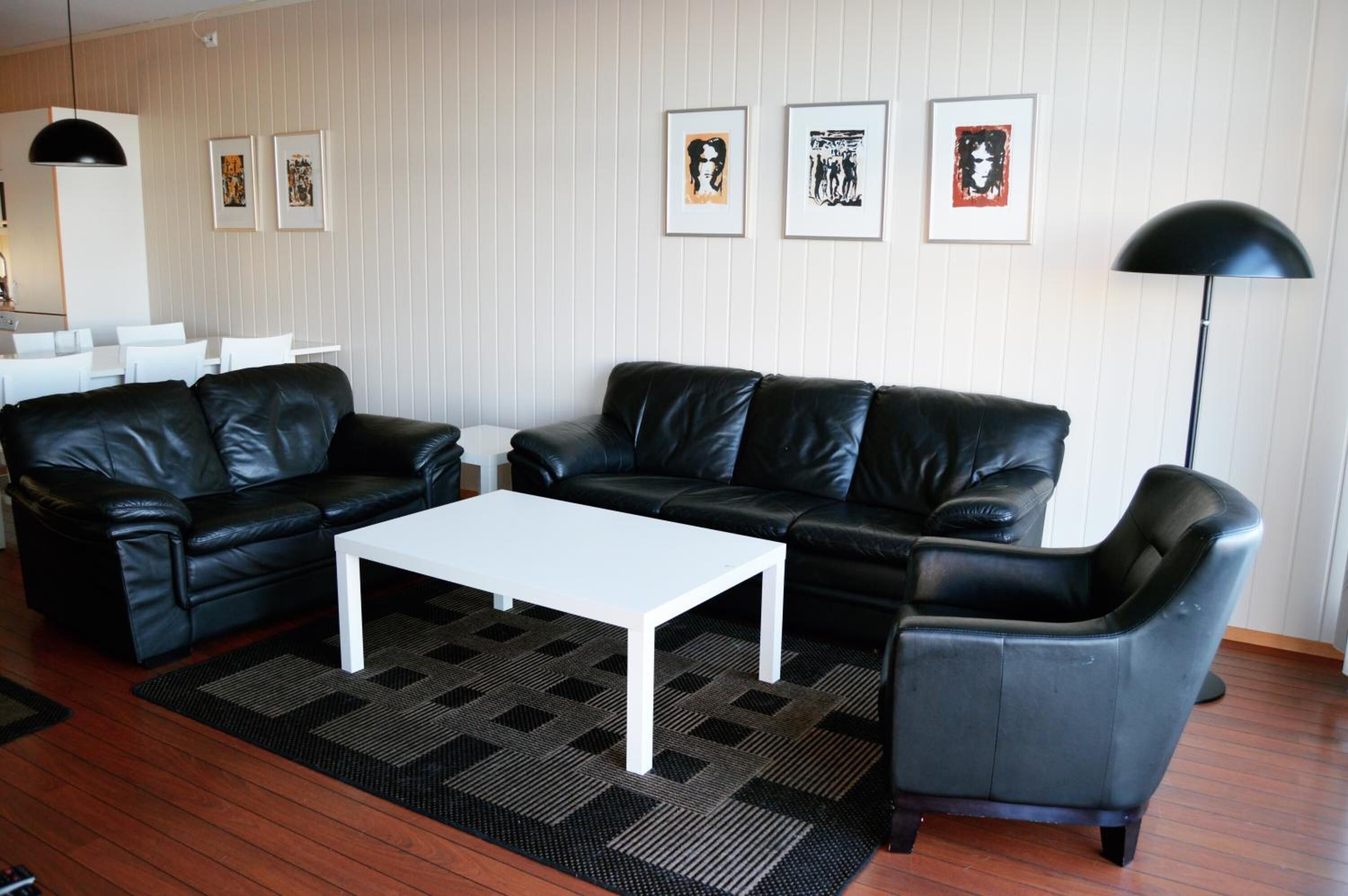 Sala de estar com sofá de couro preto, mesa de centro branca e luminária de chão preta. Quadros emoldurados na parede e área de jantar com cadeiras e mesa brancas.