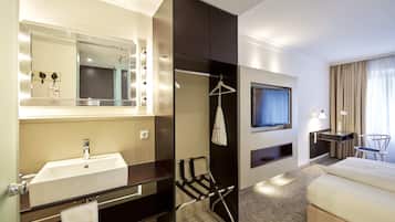 Quarto de hotel com um espelho grande, pia, porta-toalhas, televisão, cama e uma cadeira.