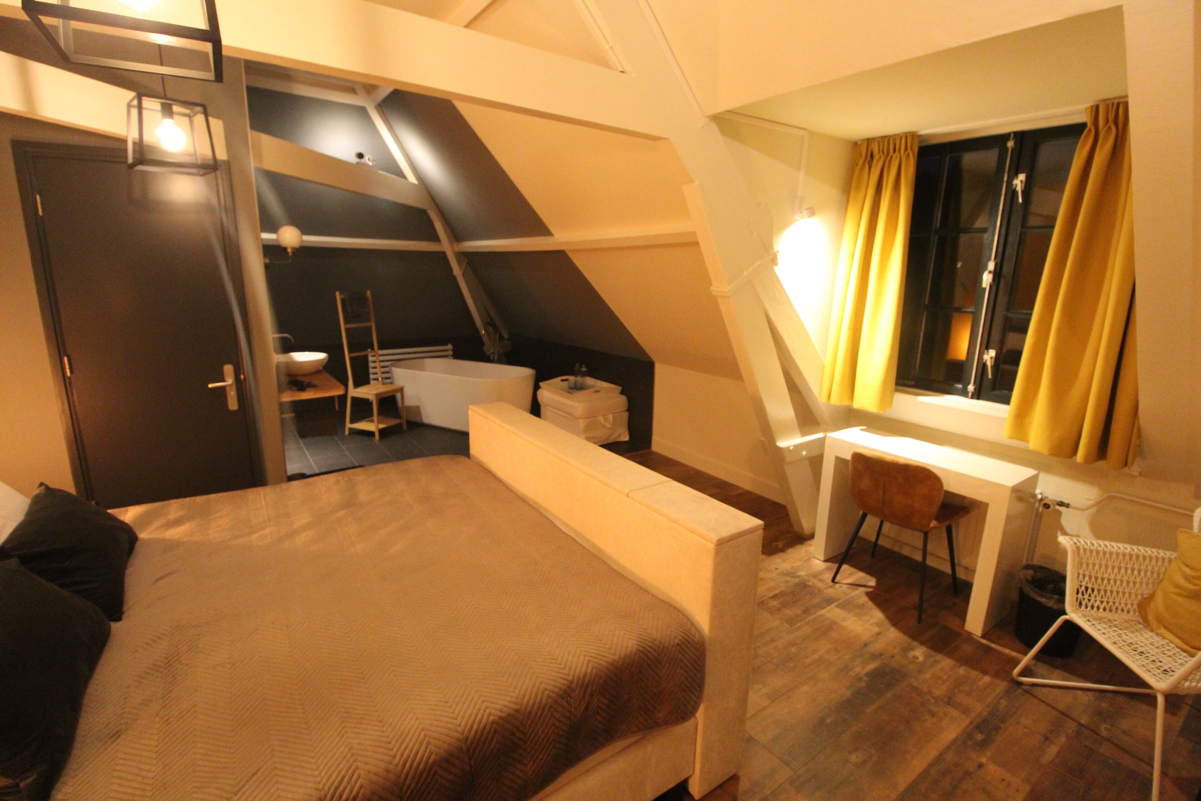 Quarto com cama, escrivaninha e cadeira. Há uma janela com cortinas, uma porta e uma mesinha de cabeceira com abajur.