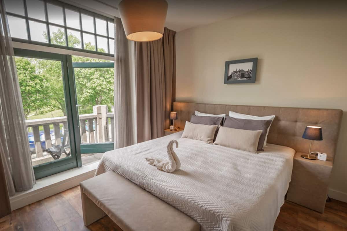 Quarto de hotel com uma cama grande, um banco, uma varanda com vista e um quadro na parede.