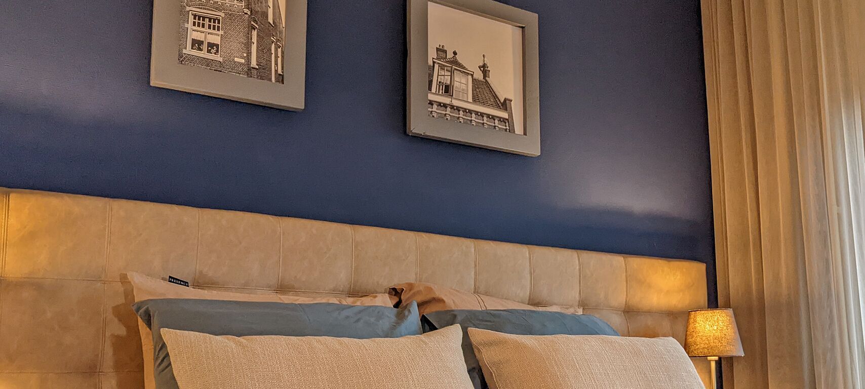 Quarto com cabeceira estofada, cama com várias almofadas e duas molduras na parede.