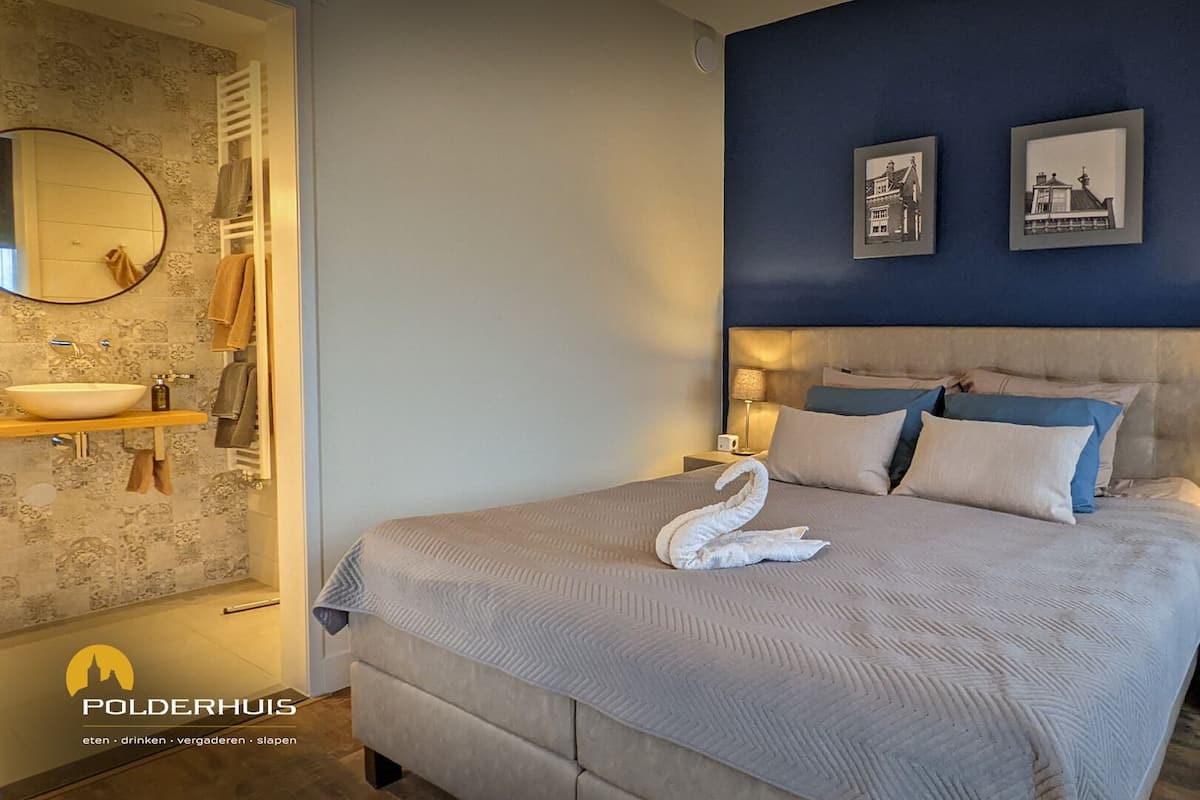 Quarto de hotel com uma cama grande, cabeceira, mesa de cabeceira com abajur, um criado-mudo, espelho, porta-toalhas e parede com duas molduras.