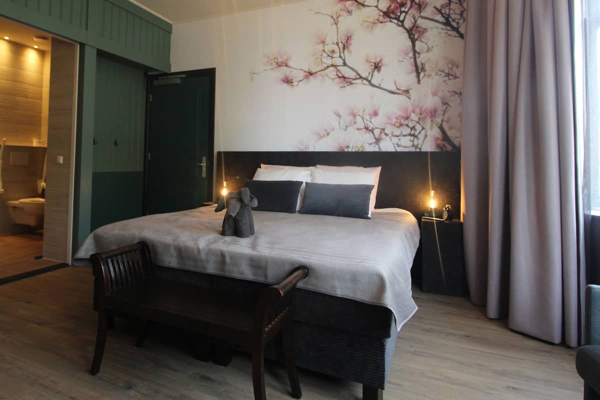 Quarto de hotel com cama, um banquinho, uma mesa de cabeceira e um banheiro visível por uma porta aberta.