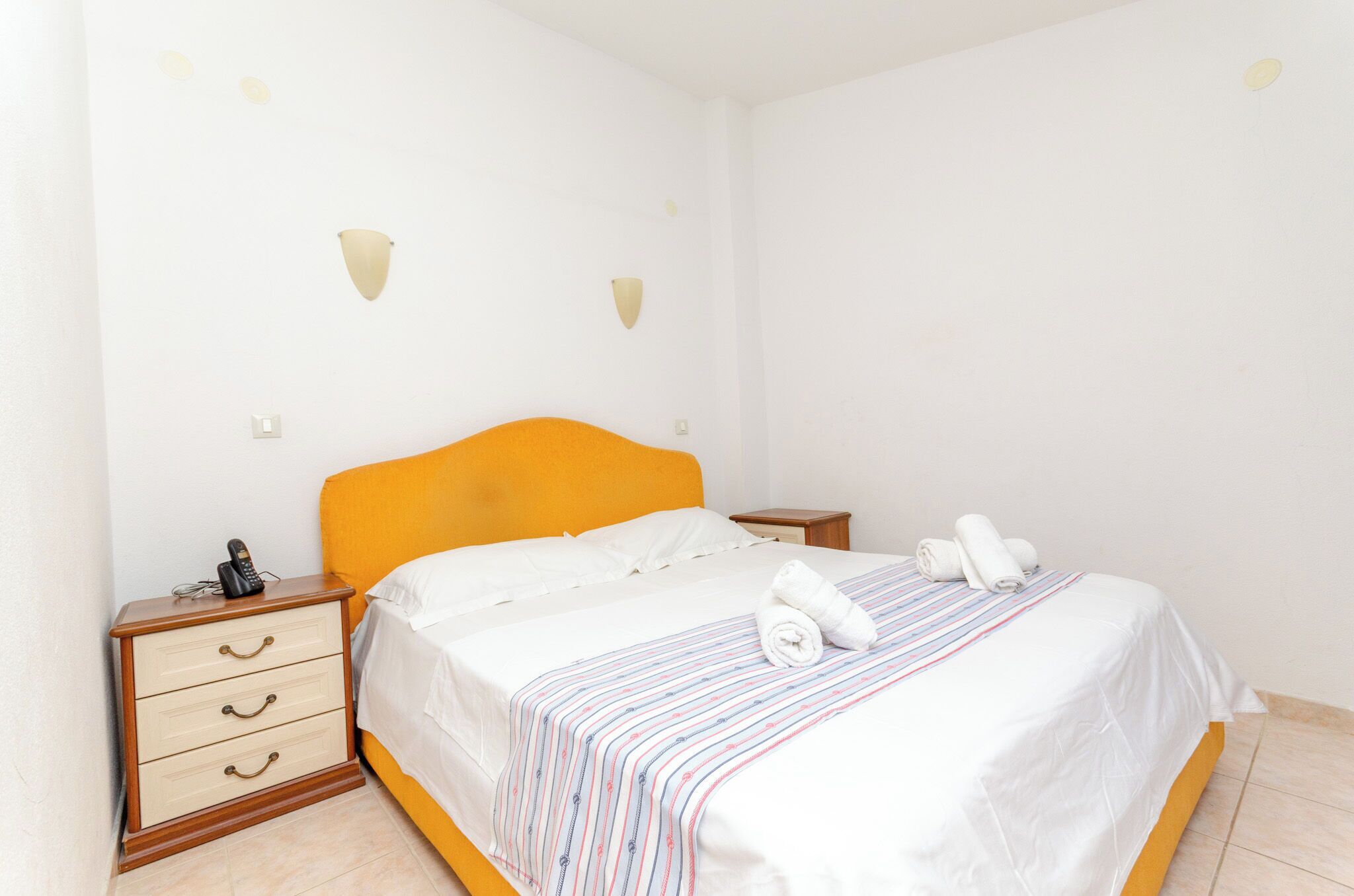 Uma cama bem arrumada com um cobertor listrado, uma mesa de cabeceira com um telefone e uma mesinha de cabeceira com gavetas.