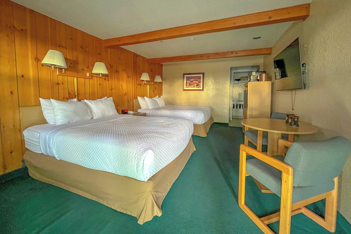 Quarto de hotel com duas camas, uma mesinha, uma cadeira, televisão e paredes de madeira.