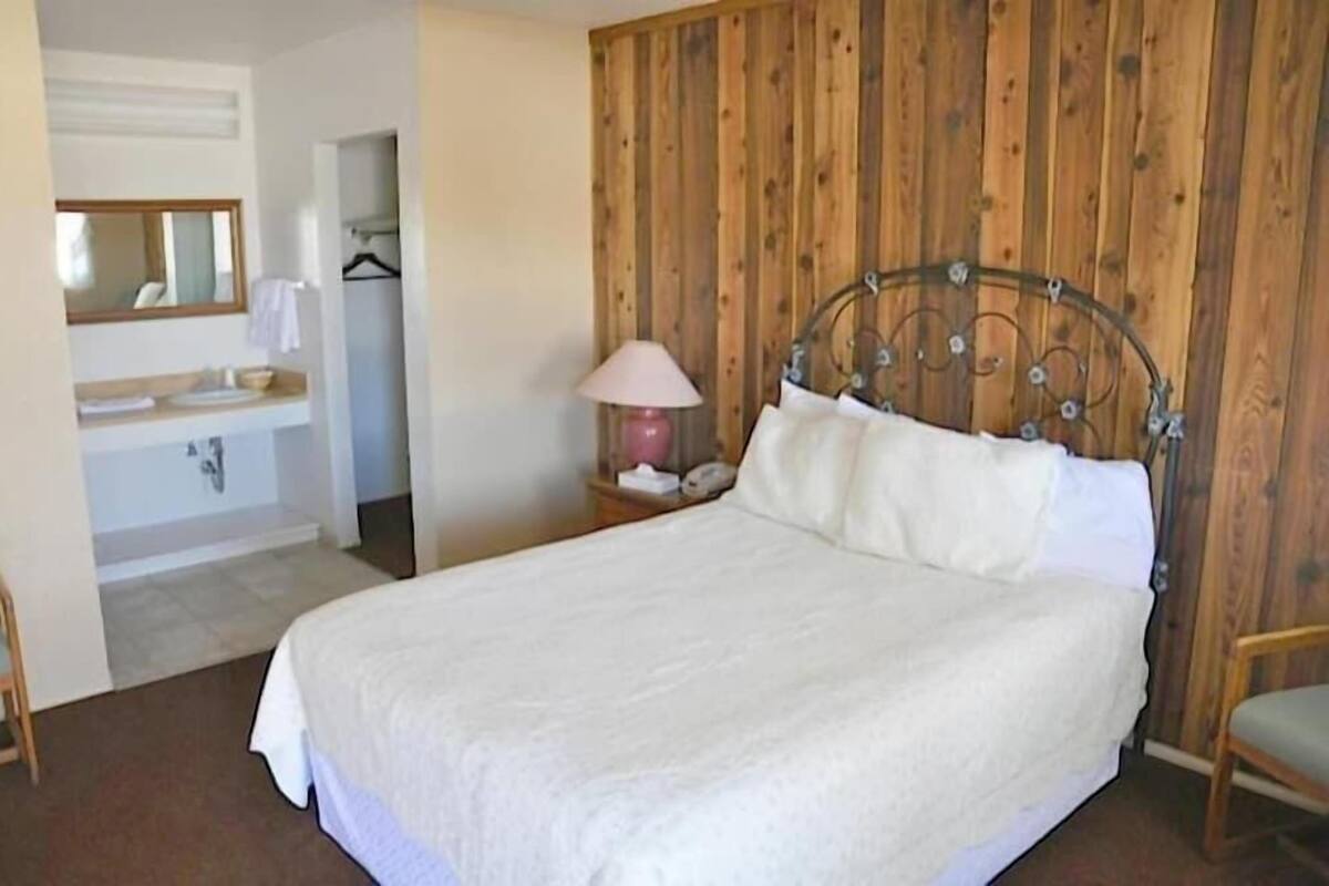 Quarto com cama, cabeceira de madeira, mesa de cabeceira com luminária e um banheiro visível ao fundo.
