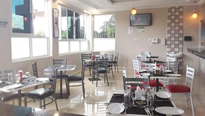 Uma sala de jantar com várias mesas e cadeiras, janelas amplas e uma televisão fixada na parede.