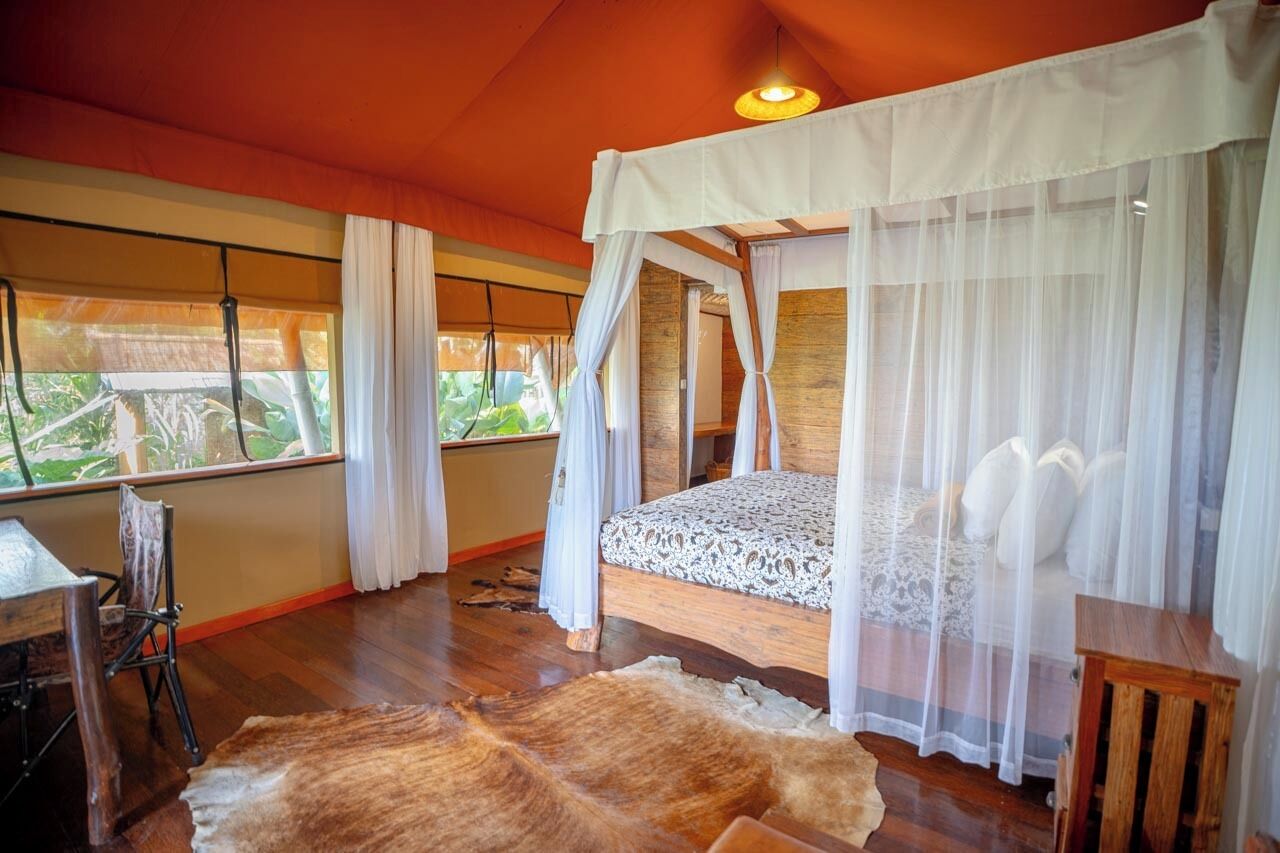 Quarto com cama de dossel, piso de madeira, escrivaninha com cadeira e vista para o verde lá fora.