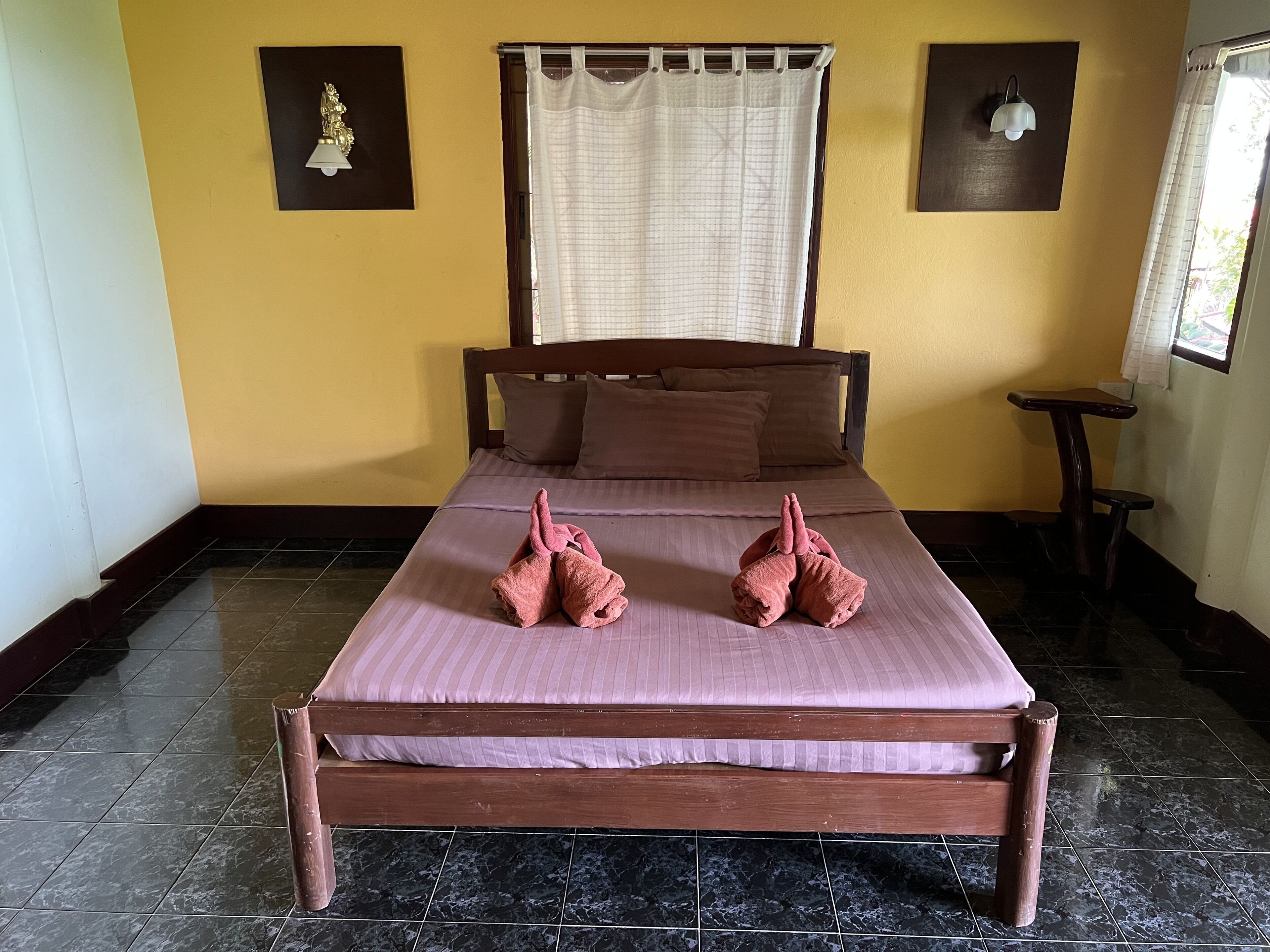 Uma cama com estrutura de madeira, uma colcha rosa e duas toalhas dobradas em posição de oração.