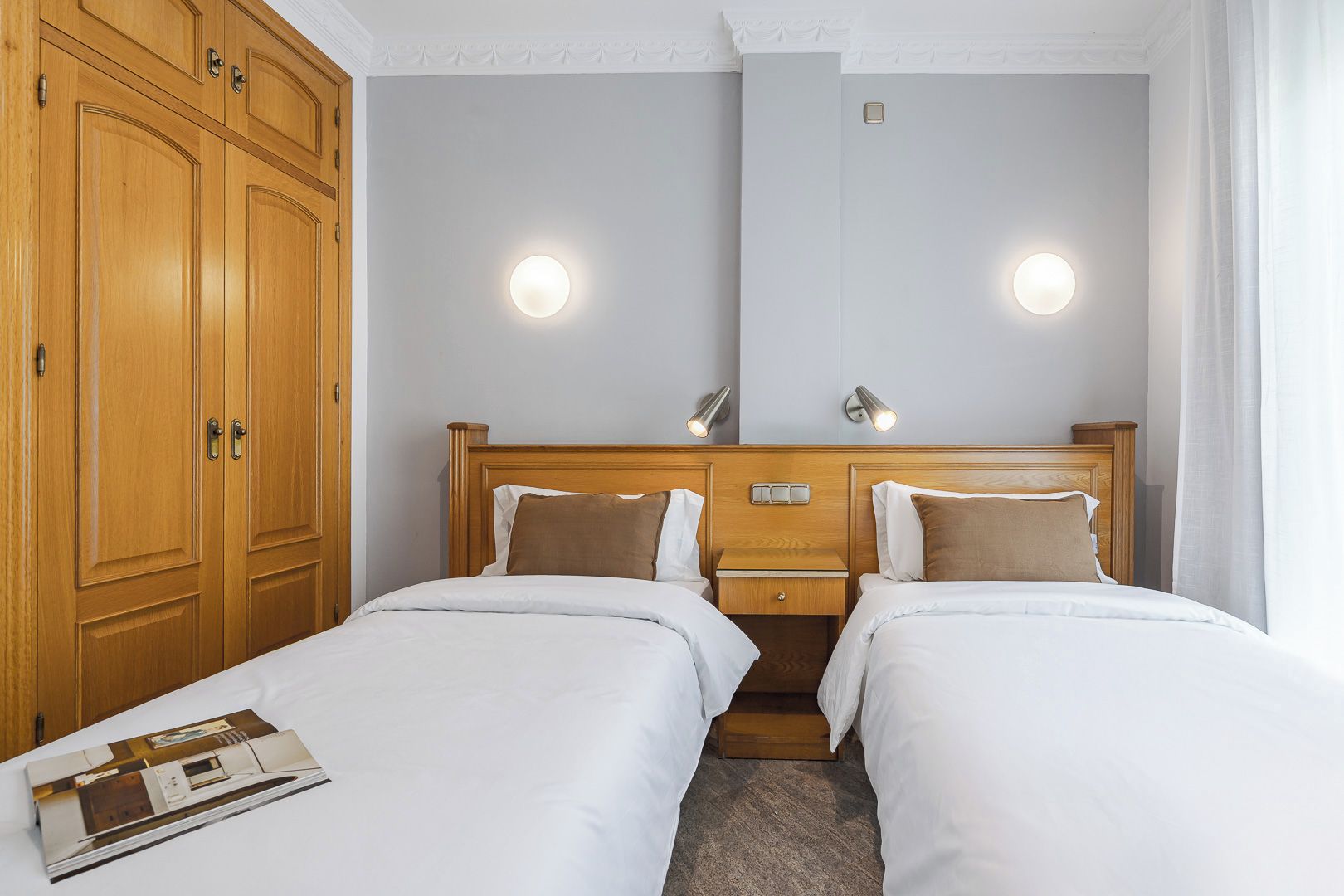 Quarto de hotel com duas camas de solteiro, guarda-roupa de madeira e luminárias de cabeceira.