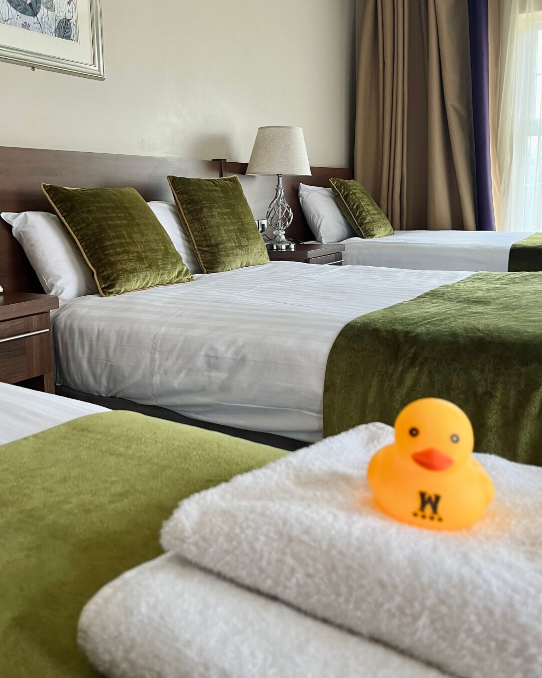 Quarto de hotel com duas camas, roupa de cama verde e branca, uma lâmpada, um quadro na parede e um patinho de borracha em cima de uma toalha.