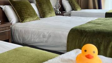 Quarto de hotel com duas camas, roupa de cama verde e branca, uma lâmpada, um quadro na parede e um patinho de borracha em cima de uma toalha.