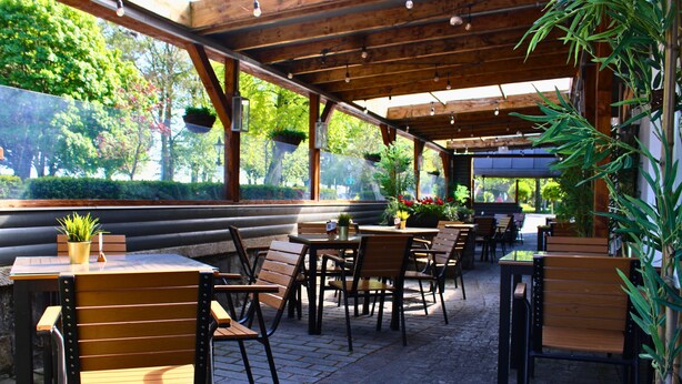 Um café ao ar livre com mesas e cadeiras de madeira, plantas em vasos e vista para o verde através de grandes janelas.