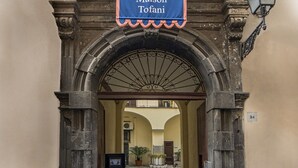 Uma entrada com portão de ferro forjado, um banner azul com o texto "Maison Tofani" e um arco de pedra.