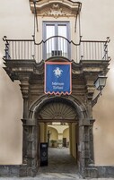 Uma entrada com portão de ferro forjado, um banner azul com o texto "Maison Tofani" e um arco de pedra.