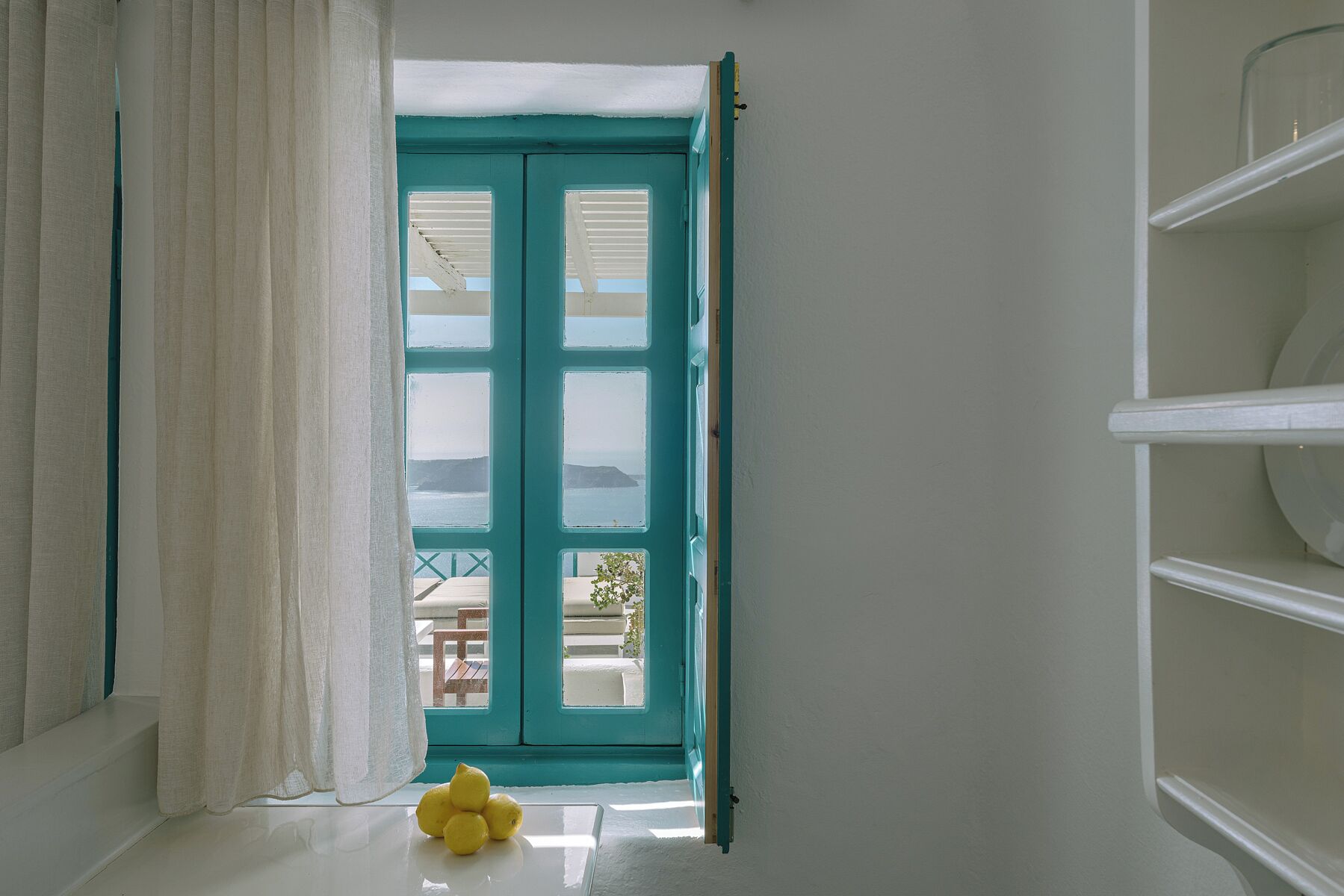 Um quarto com janela emoldurada em azul-esverdeado, uma prateleira branca e uma cortina branca.