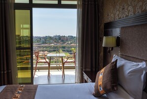 Quarto de hotel com uma cama, uma mesa de cabeceira com abajur, e vista da cidade através de uma varanda com mesa e cadeiras.