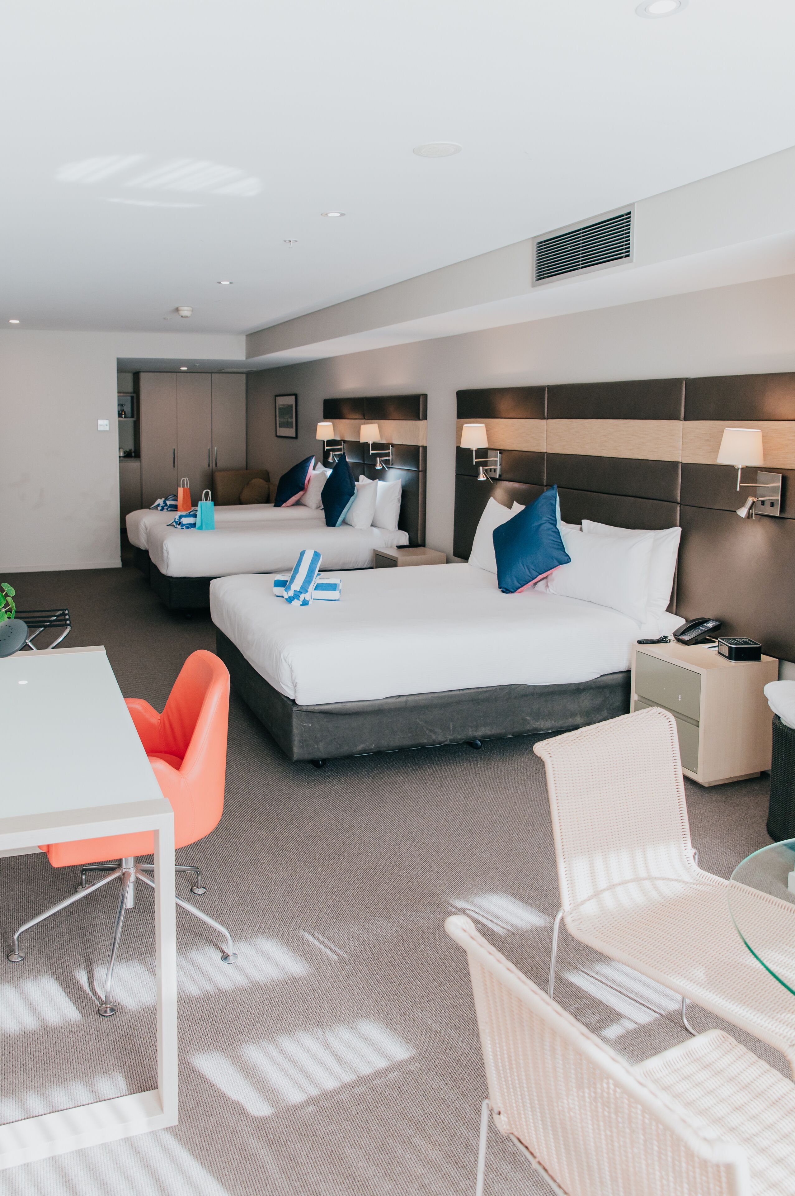 Quarto de hotel com duas camas, uma escrivaninha, uma cadeira e uma mesinha.