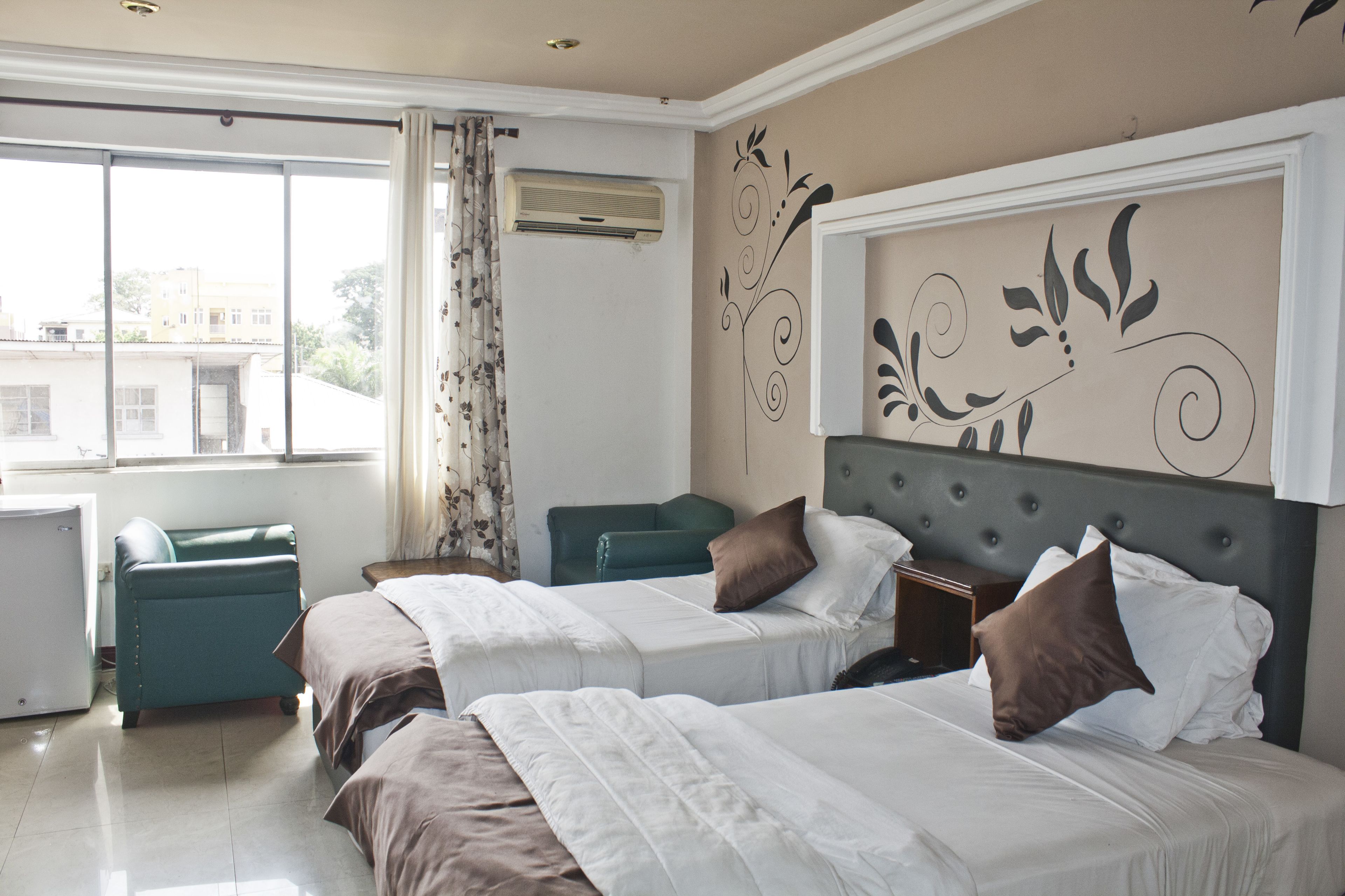 Quarto de hotel com duas camas, uma poltrona verde, uma mesinha e uma janela com cortinas.