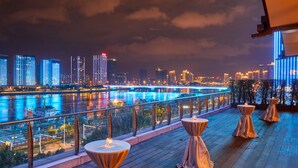 Vista noturna de um restaurante à beira-rio, com mesas e cadeiras, com vista para um rio e luzes da cidade ao fundo.