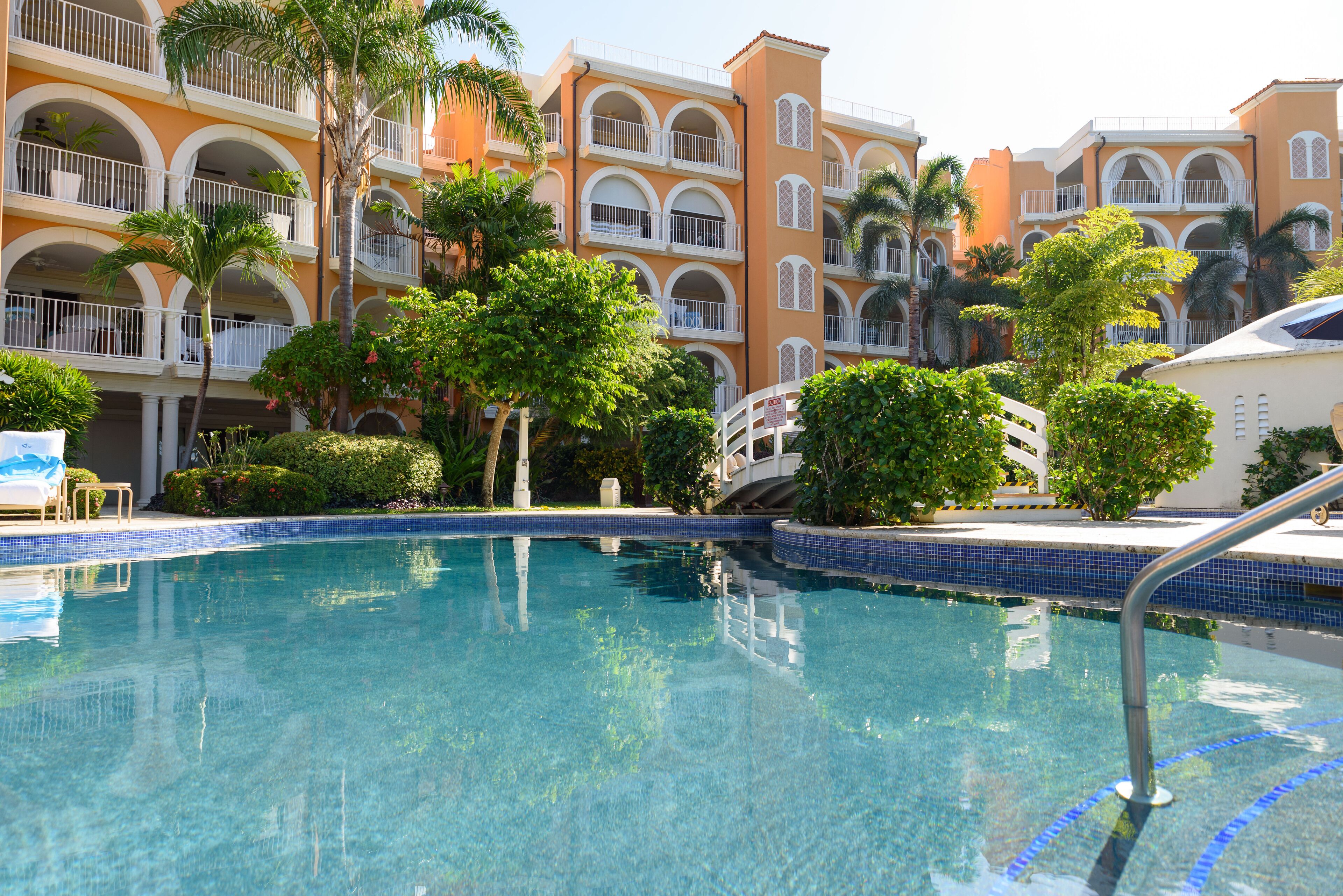 Piscina com água azul cristalina, rodeada por um hotel com várias andares, varandas e palmeiras.