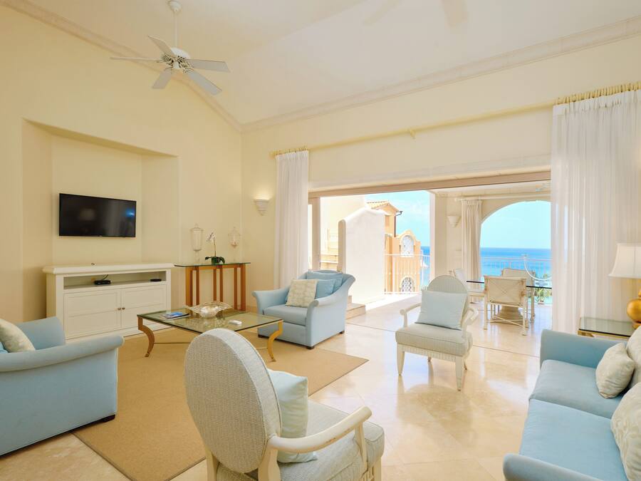 Uma sala de estar ampla, com sofás azul-claros, uma mesa de centro e uma televisão. Possui uma varanda com vista para o mar.