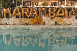 Área de piscina com balões dourados e brancos formando a palavra 'NARAIBA', com reflexo dos balões e da piscina na água.