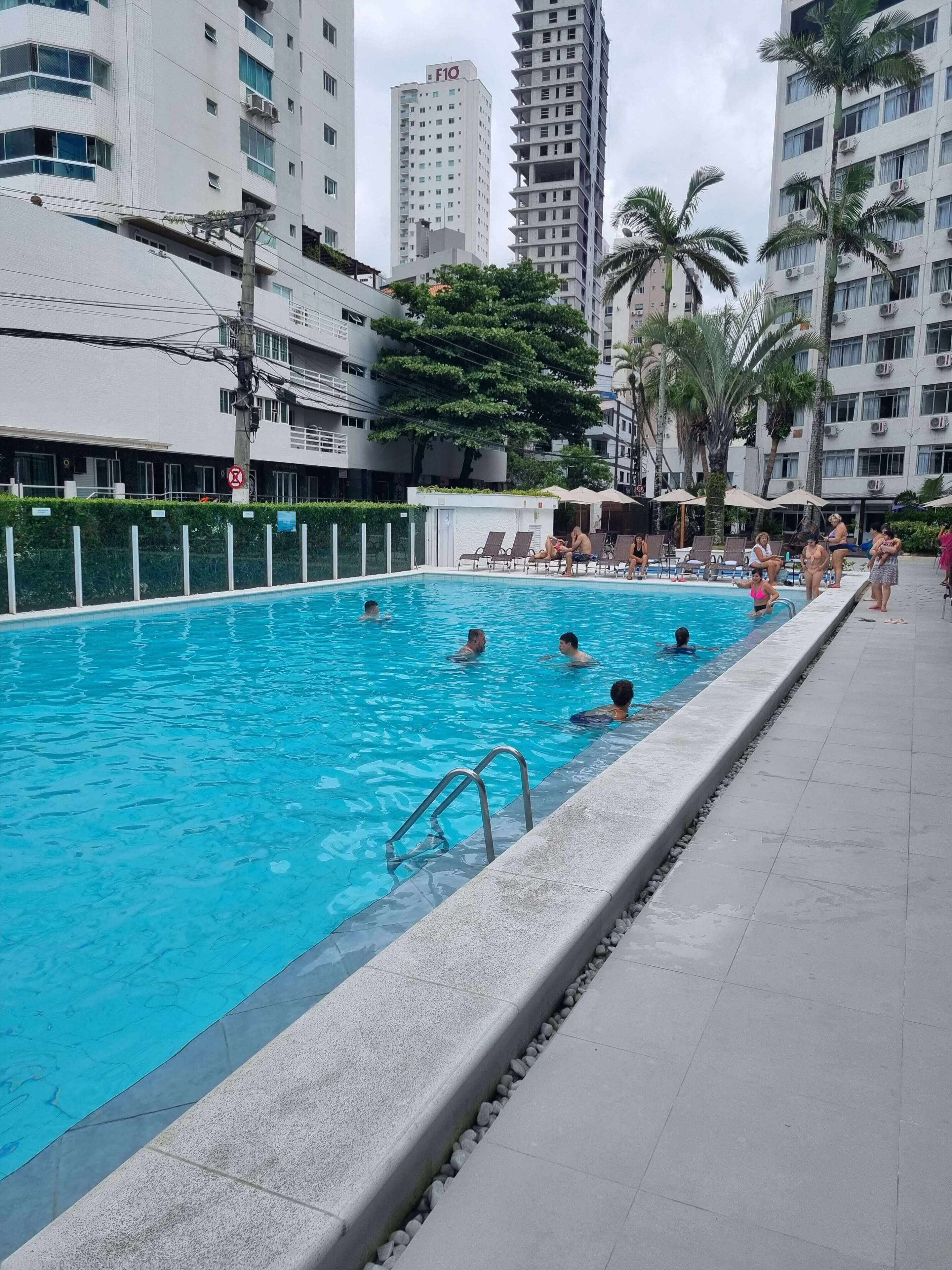 Uma piscina com pessoas nadando e tomando sol, cercada por palmeiras e edifícios modernos.