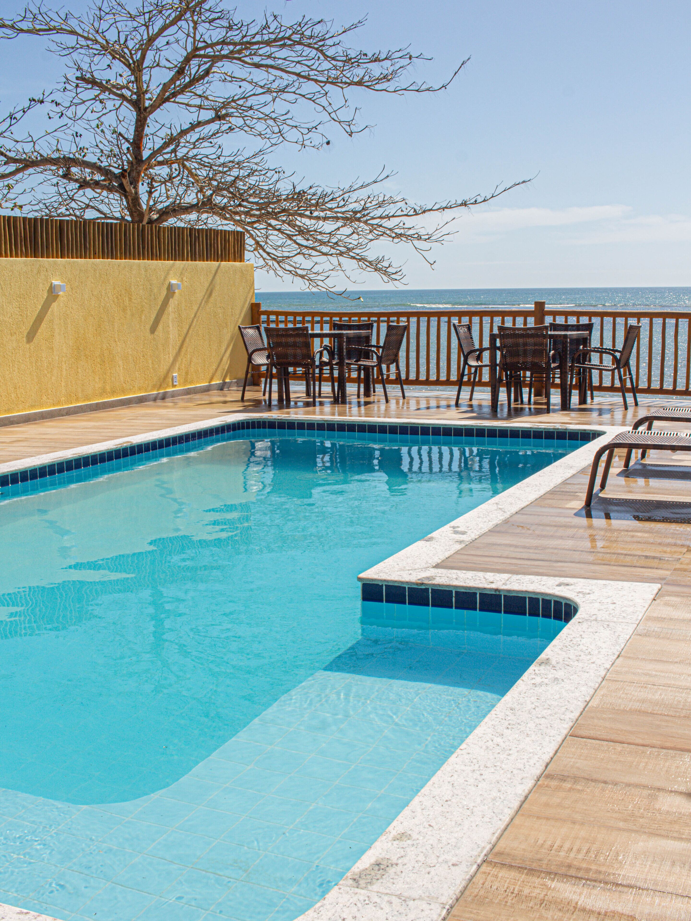 Piscina com deck de madeira, rodeada por cadeiras e mesas de vime, com vista para um corpo d'água.