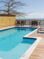Piscina com deck de madeira, rodeada por cadeiras e mesas de vime, com vista para um corpo d'água.