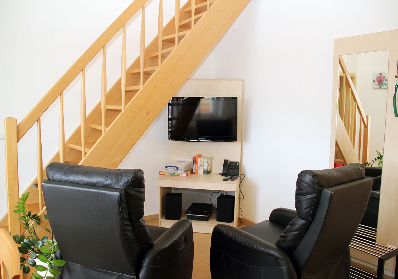 Uma sala de estar com escada de madeira, televisão de tela plana, duas poltronas de couro preto, uma mesa pequena com livros e um telefone, e uma planta.