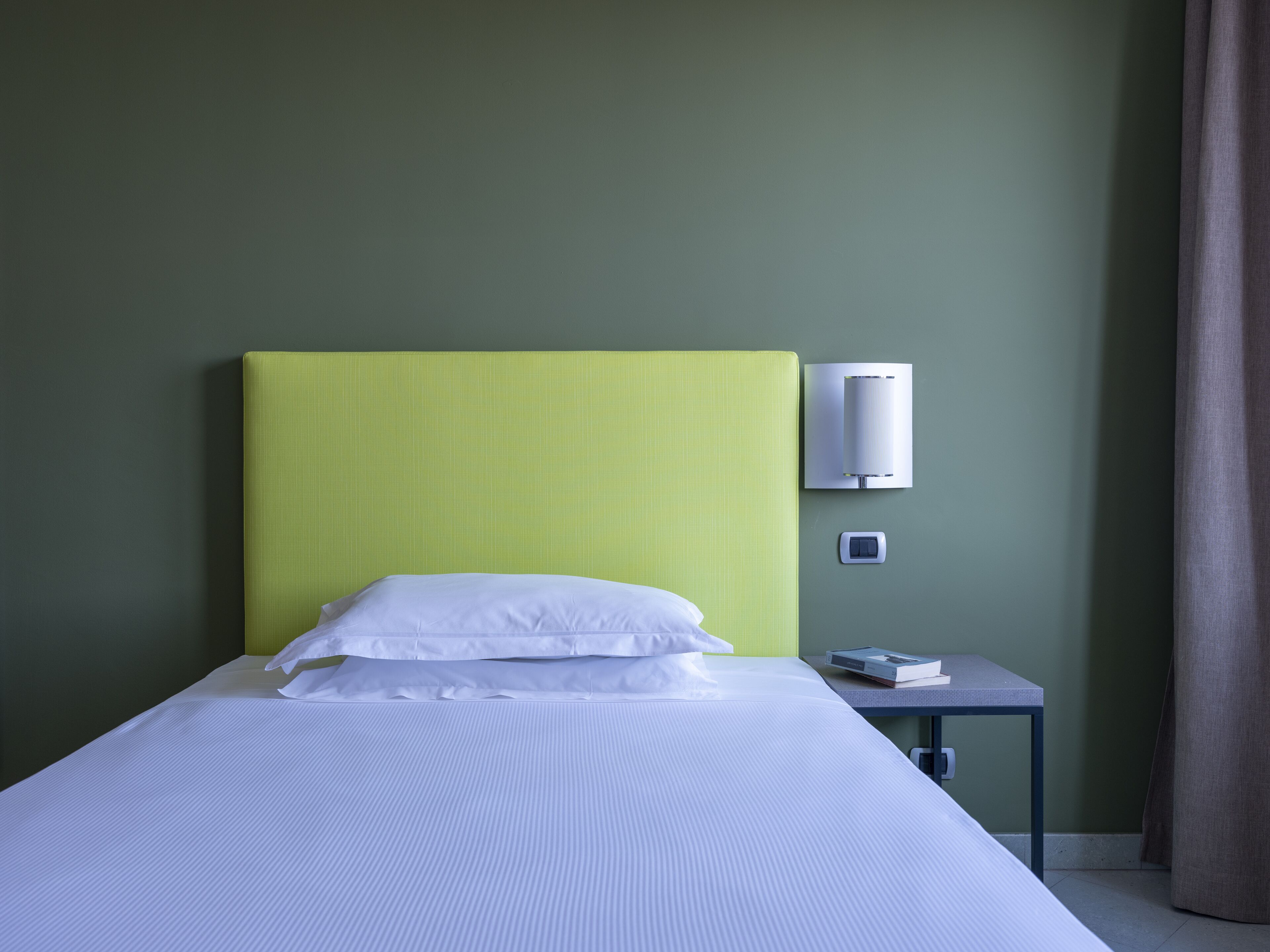 Quarto de hotel com cabeceira verde, roupa de cama branca, mesa de cabeceira com um livro e uma luminária na parede.
